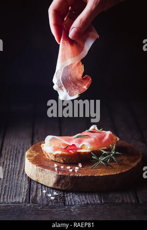 Female hand putting prosciutto on bread over dark wooden background ...