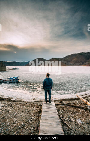 Back view of man standing on shore of lake with cliffs on background ...