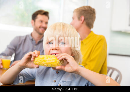 Boy eating corn cob in kitchen with parents in background Stock Photo ...