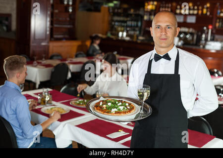 Polite waiter holding tray in restaurant with customers behind him ...