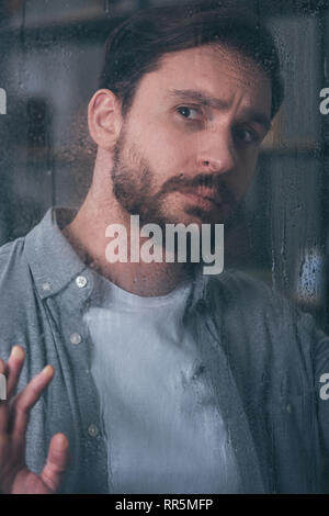 handsome upset man touching window with raindrops and looking away ...