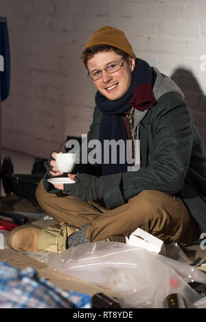 smiling homeless man drinking coffee while sitting by white brick wall ...