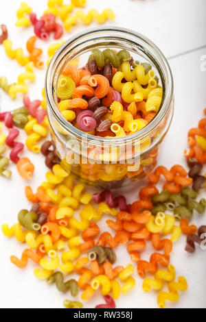 uncooked multi-colored pasta poured from a jar on a white table. space ...