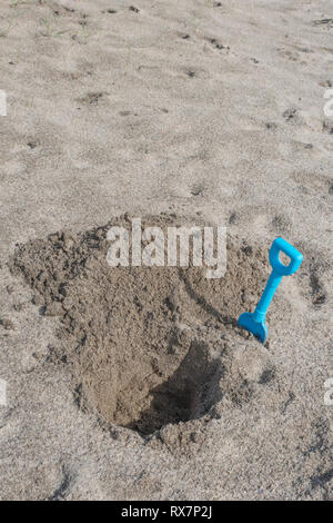 Seaside hole dug into sand, with kids blue spade. Metaphor 'dig a hole ...