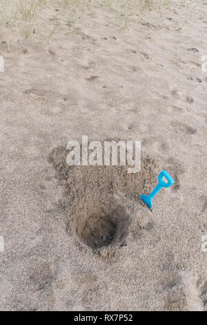 Seaside hole dug into sand, with kids blue spade. Metaphor 'dig a hole ...