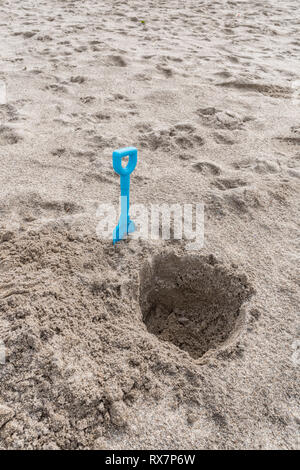Seaside hole dug into sand, with kids blue spade. Metaphor 'dig a hole ...