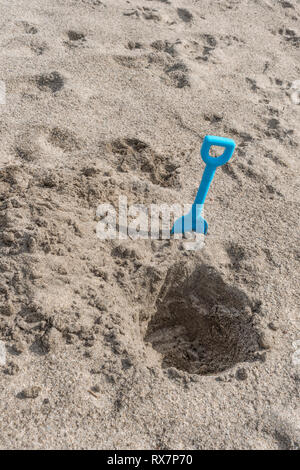 Seaside hole dug into sand, with kids blue spade. Metaphor 'dig a hole ...