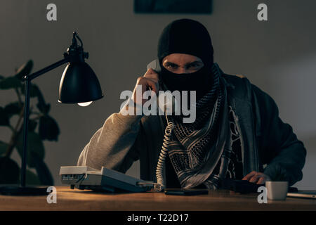Angry terrorist in mask talking on telephone and showing gun at webcam ...