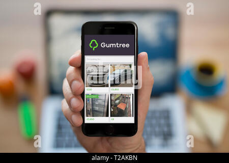 A man looks at his iPhone which displays the Gumtree logo, while sat at ...