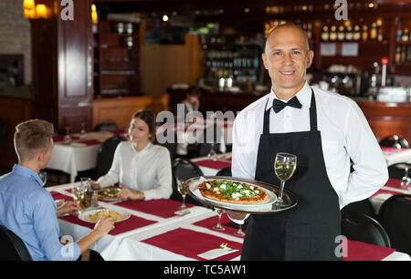 Polite waiter holding tray in restaurant with customers behind him ...