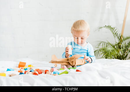 toddler boy playing with wooden biplane near colorful toy blocks on bed ...