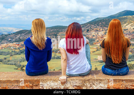 A group of three girls, with their backs to the camera, sit on a bench ...