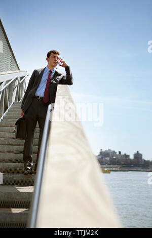 Businessman leaning against railing, briefcase on sidewalk at feet ...