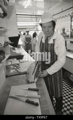 1980s, a traditional butcher in a whitecoat, apron and hat in his ...
