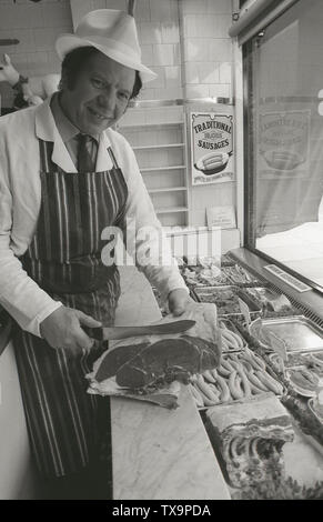 1980s, a traditional butcher in a whitecoat, apron and hat at the back ...