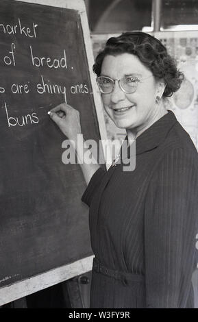 1950s, historica, a smiling lady teacher writing with chalk at her ...