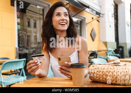 amazed woman holding paper cup and using smartphone isolated on pink ...