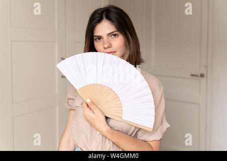 Brunette woman holding up fan in front of face, smiling, looking away ...