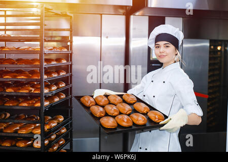 Girl baker holds a tray with hot pastries in the bakery. She is wearing ...