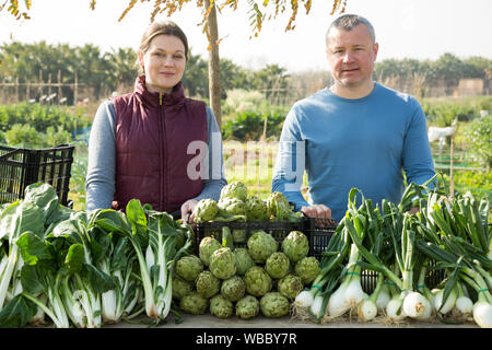 Cheerful couple of farmers standing behind counter with fresh greens ...