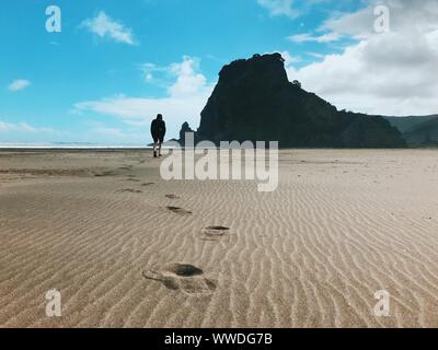 Man walking towards the Lion Rock at Piha Beach, blue sunny day ...