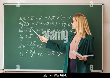 Beautiful math teacher with book and pointer near blackboard in ...