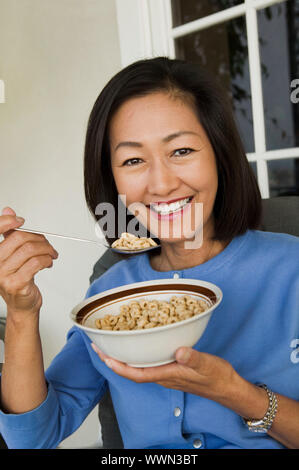 Asian women eating cereal breakfast bowl sitting on living room couch ...