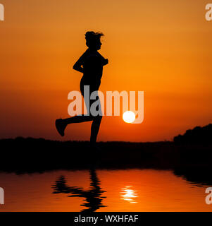 Silhouette woman running against orange sunset with reflection in water ...