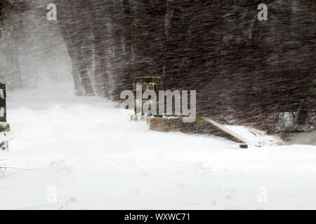 Snow blowing sideways over a pond and a bridge with a park bench on the ...