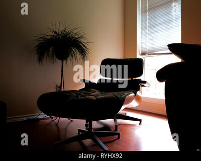 Charles und Ray Eames Designer schwarz Lounge Sessel mit Ottomane in einem Wohnzimmer Einstellung durch ein Fenster auf einen Holzboden gedampften Tonen und dunkel Stockfotografie Alamy